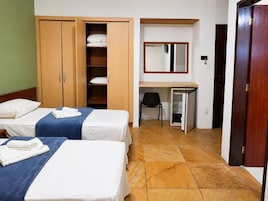 Quarto de hotel com duas camas, um guarda-roupa, uma secretária com cadeira e um espelho.