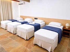 Quarto de hotel com quatro camas de solteiro dispostas em linha, cabeceira de madeira, mesa de cabeceira com abajur e relógio digital.