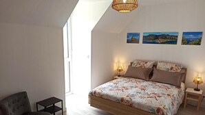 Quarto com cama, uma cadeira, uma mesinha e um aquecedor. Há três quadros na parede.