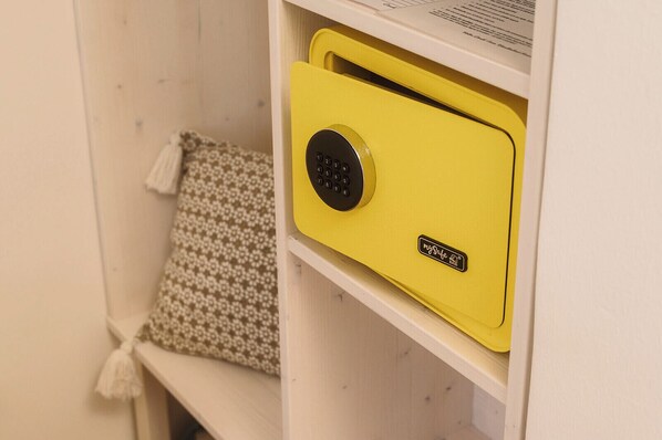 Um cofre amarelo com uma fechadura e o logotipo 'mySafe', um travesseiro estampado e uma toalha branca numa prateleira.
