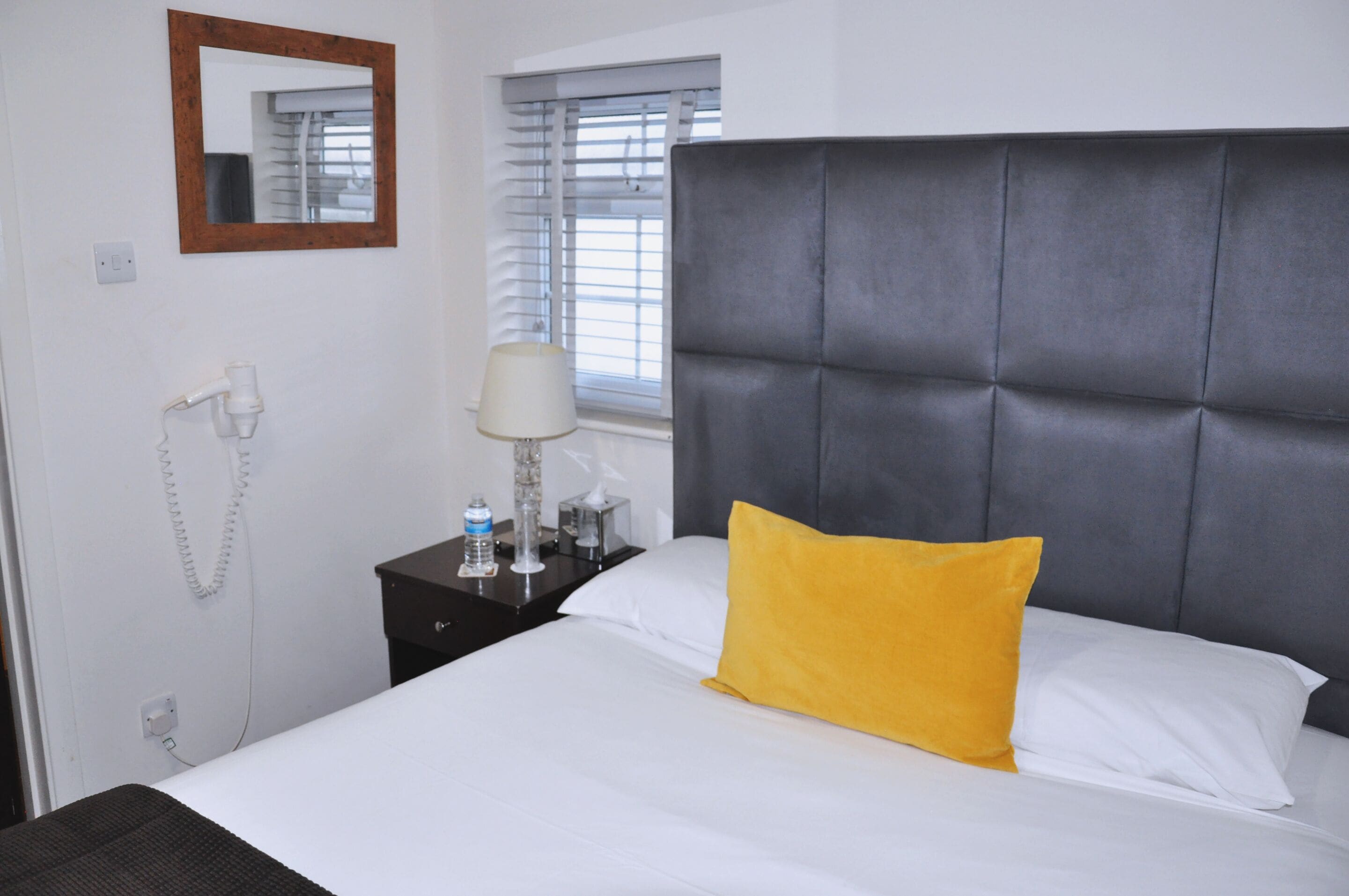 Quarto de hotel com cama, mesa de cabeceira, luminária, espelho e janela com persianas.