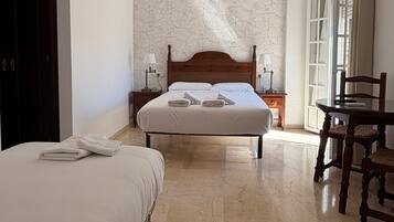 Quarto de hotel com uma cama grande, uma cama menor, uma cadeira de madeira, uma mesa e uma mesinha de cabeceira com luminárias.