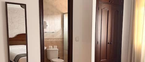 Quarto de hotel com uma cama, um banco de madeira, um banheiro com vaso sanitário e pia, e um espelho grande.