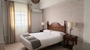 Quarto de hotel com uma cama, duas mesas de cabeceira com luminárias, uma janela com cortinas, um ar condicionado e uma parede com padrão marmorizado.