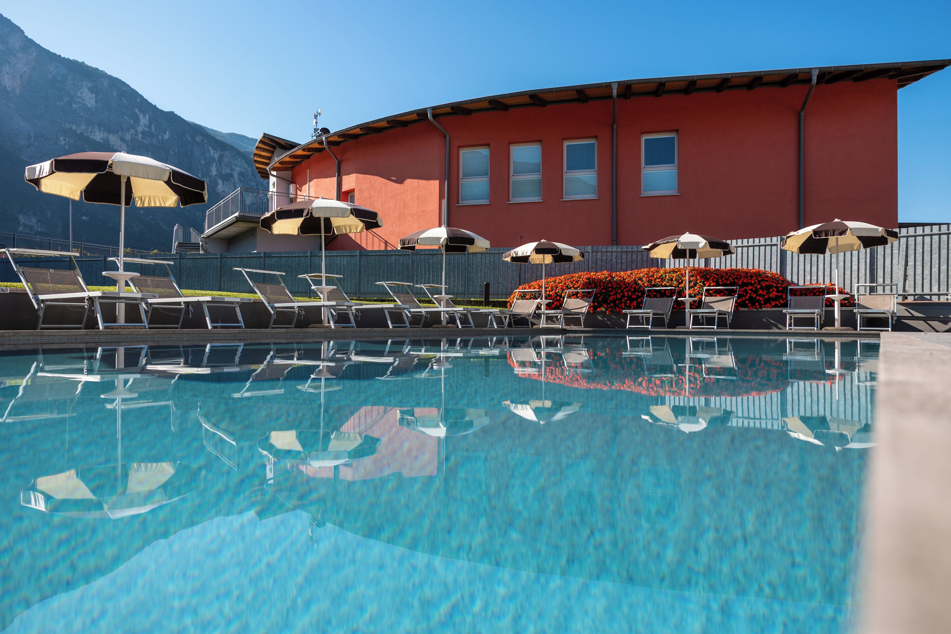 Uma piscina com espreguiçadeiras e guarda-sóis, um edifício vermelho com janelas e uma montanha ao fundo.