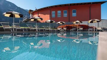 Uma piscina com espreguiçadeiras e guarda-sóis, um edifício vermelho com janelas e uma montanha ao fundo.