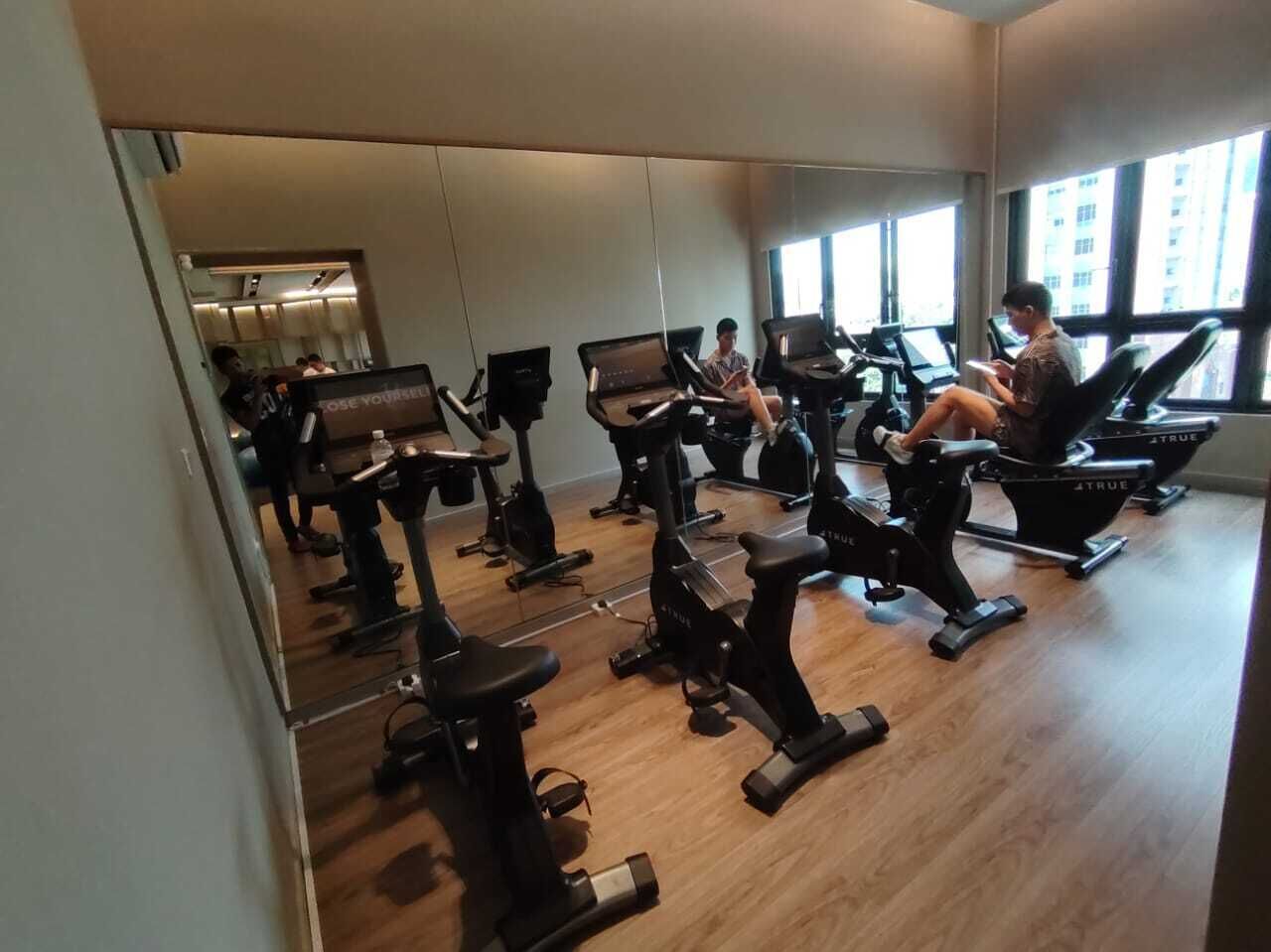 Uma academia com diversos equipamentos de exercício, janelas amplas e um espelho refletindo o ambiente.