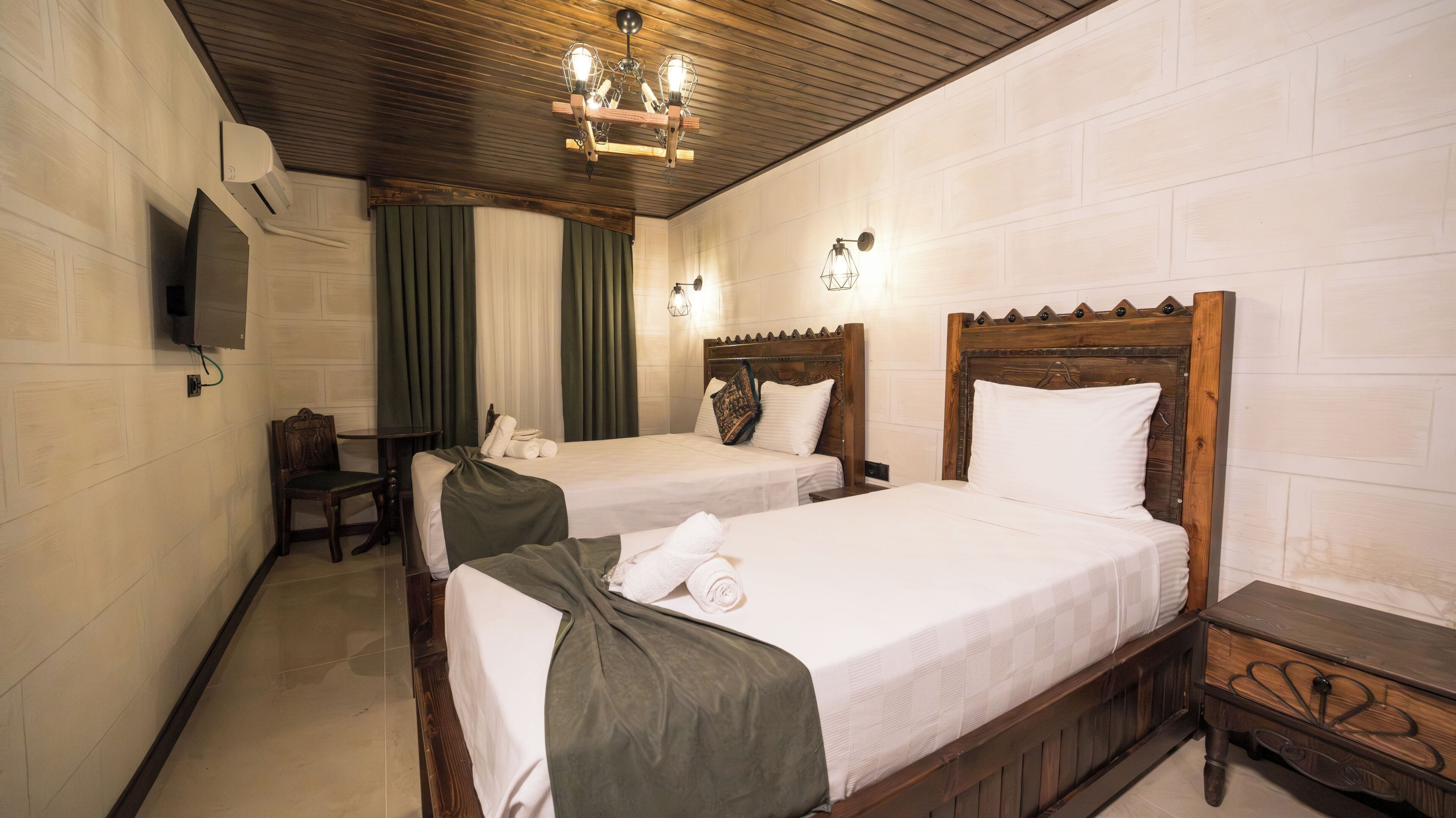 Quarto de hotel com duas camas, cabeceira de madeira, televisão fixada na parede e um candelabro.