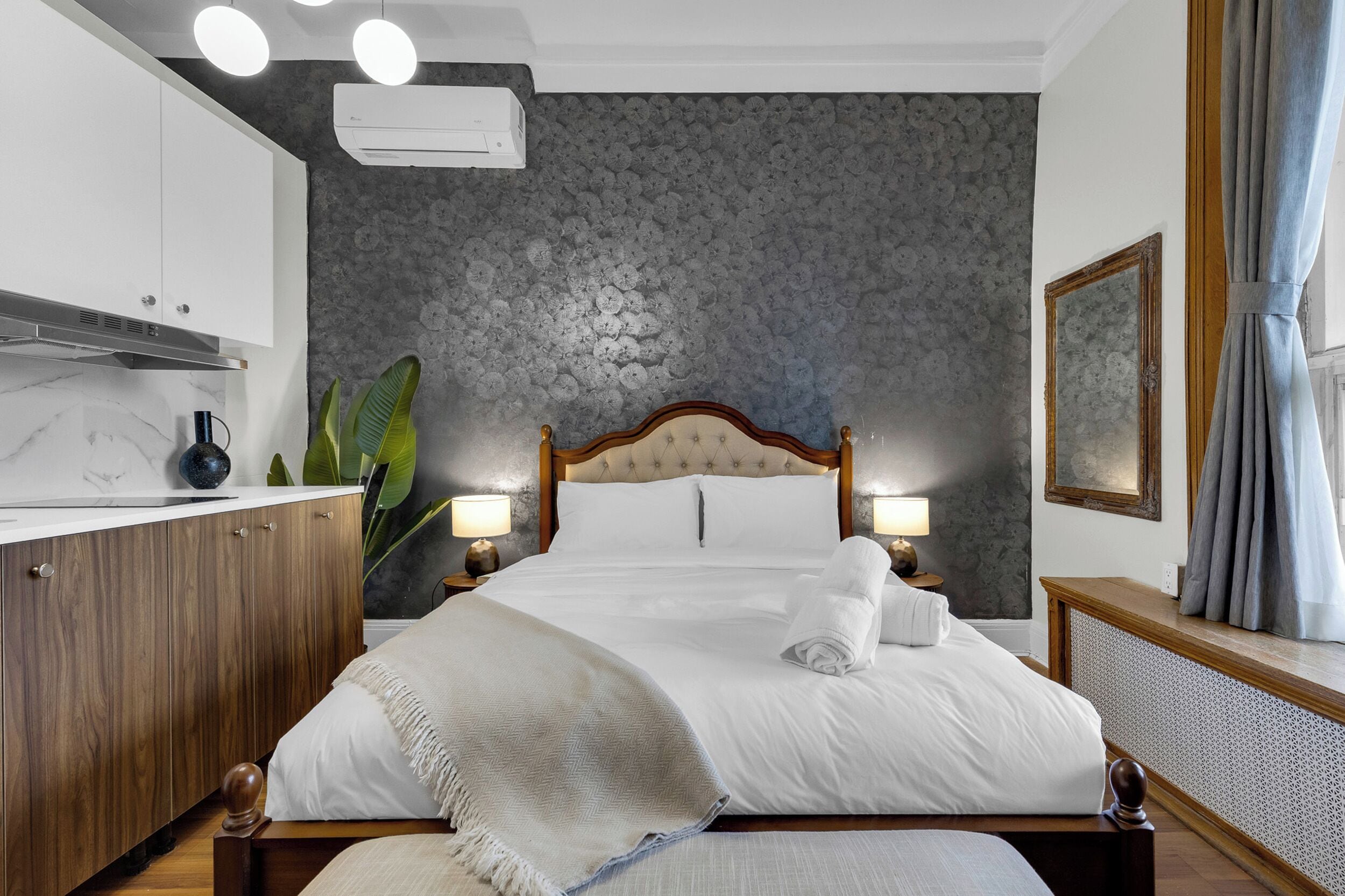 Quarto de hotel bem arrumado com cama, abajures, mesa de cabeceira de madeira, espelho e uma grande janela com cortinas.