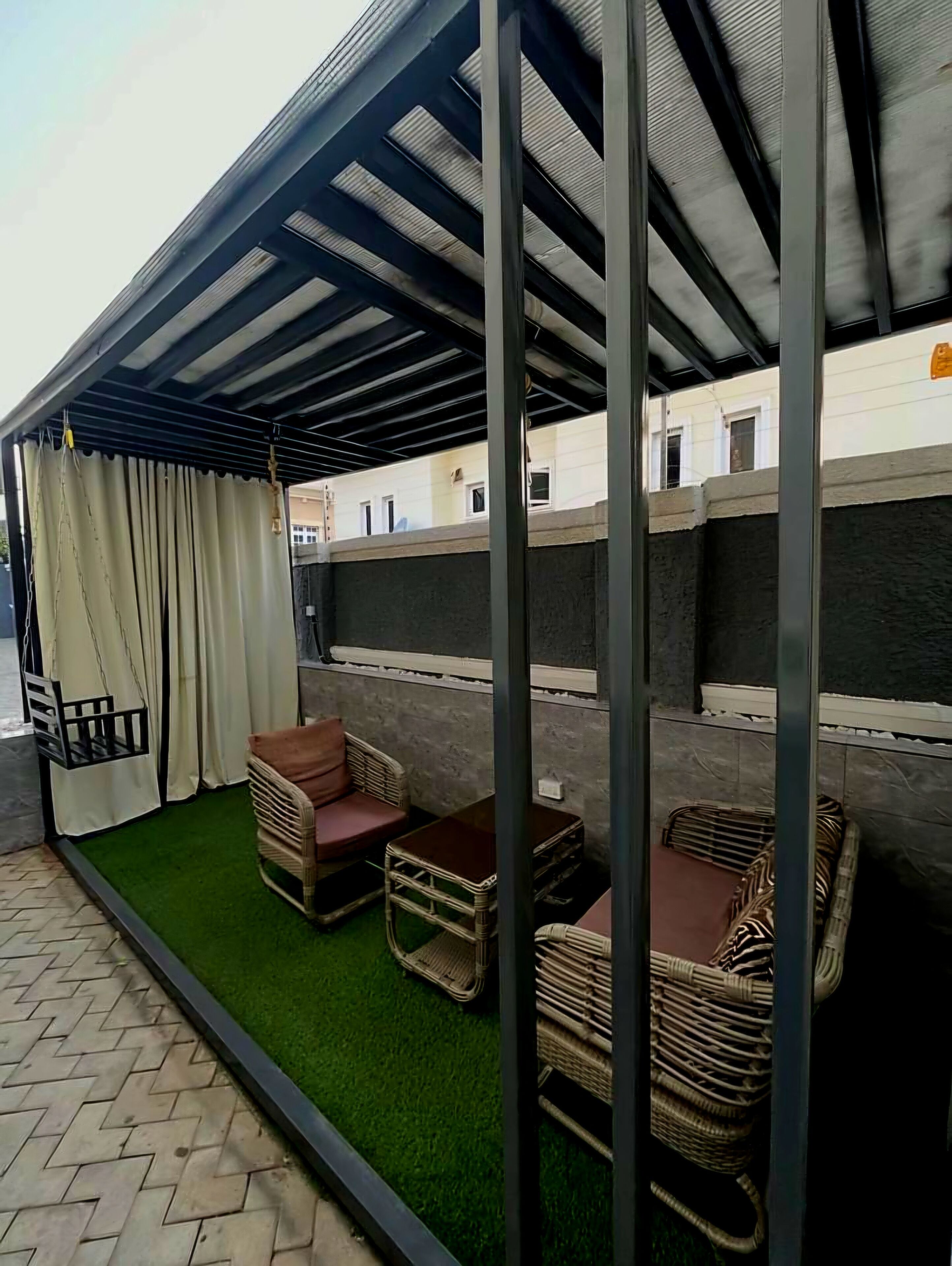 Uma área de estar externa com móveis de vime, parte coberta e vista para um edifício ao fundo.