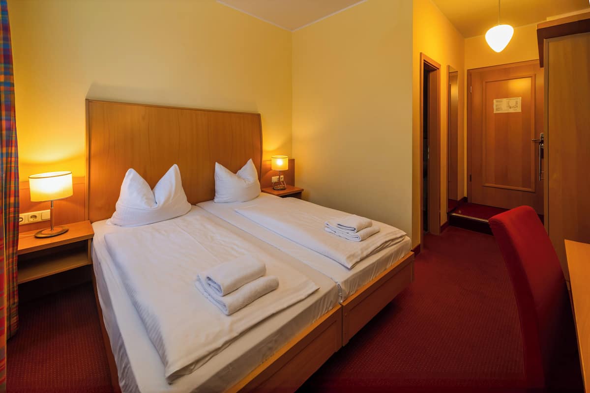 Quarto de hotel com duas camas, uma cabeceira de madeira, abajures, um tapete vermelho e uma porta.