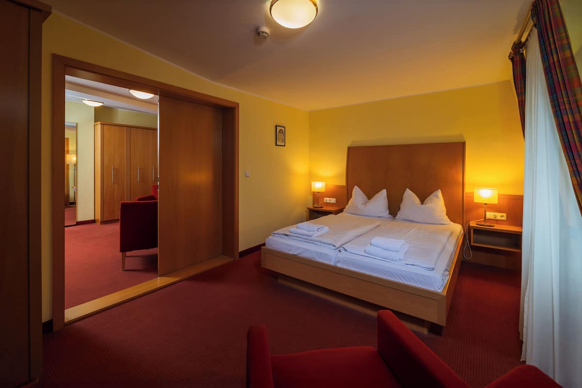 Quarto de hotel com cama, duas mesas de cabeceira, uma cadeira vermelha e uma porta que dá para outro quarto.