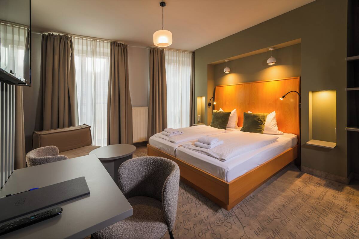 Quarto de hotel com cama, escrivaninha, cadeira, luminária e cortinas.