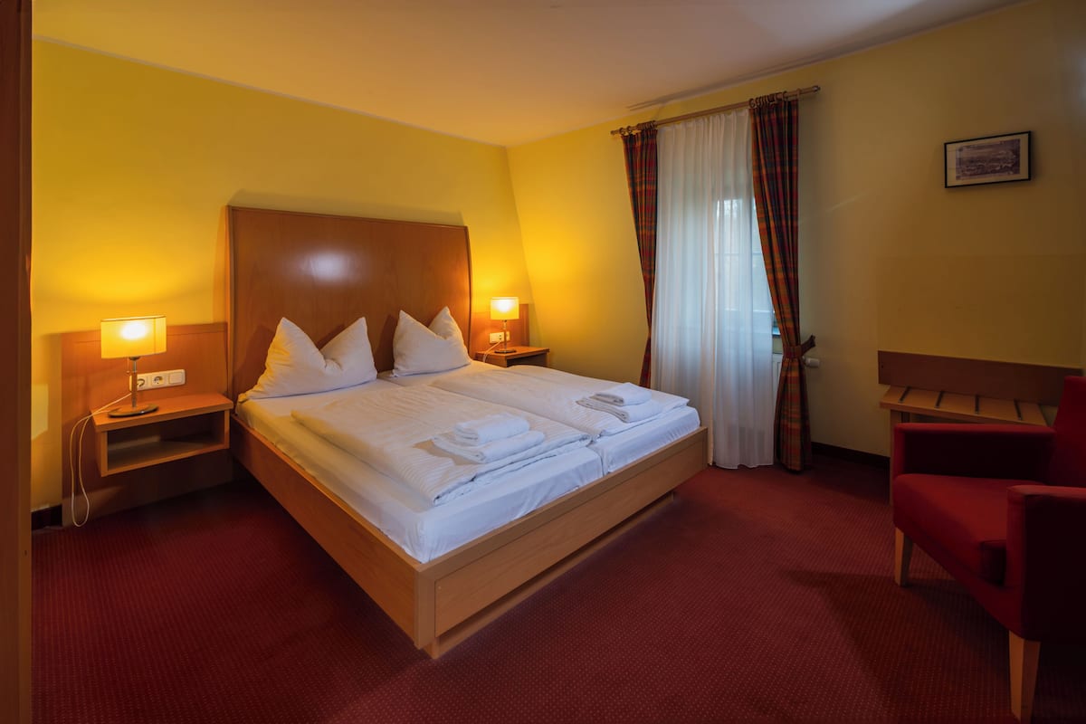 Quarto de hotel com uma cama, duas mesas de cabeceira com luminárias, uma cadeira vermelha e uma janela com cortinas.
