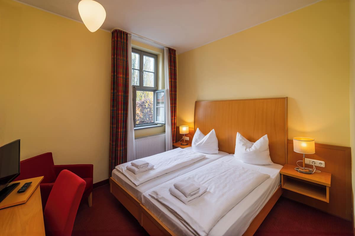 Quarto de hotel com uma cama de casal, duas mesas de cabeceira com luminárias, uma televisão e uma cadeira vermelha.