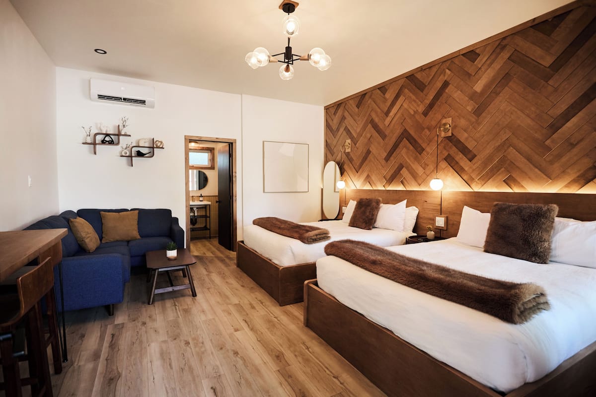 Quarto de hotel com teto em padrão herringbone de madeira, um sofá, duas camas com roupa de cama branca e almofadas marrons, uma mesa de centro de madeira e uma pequena mesa lateral.