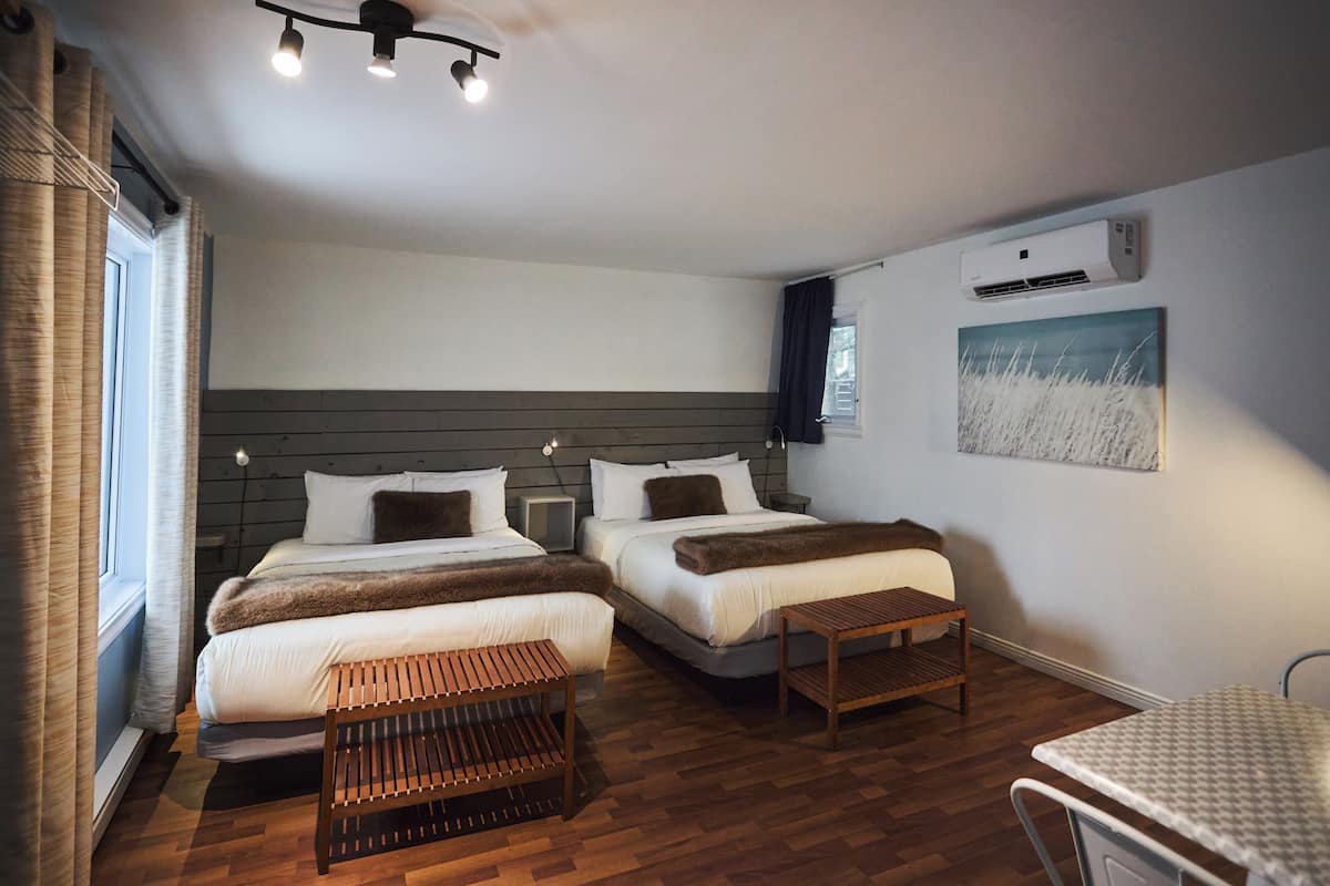 Quarto de hotel com duas camas, um banco de madeira, uma mesa e um aparelho de ar condicionado.