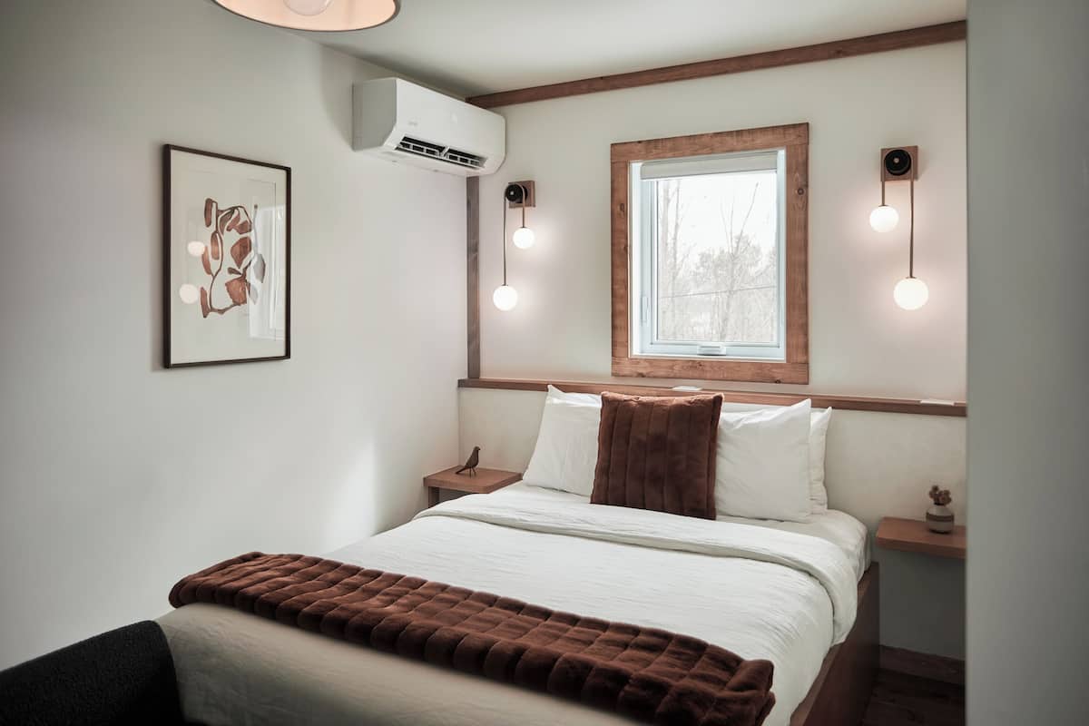 Quarto com cama, mesas de cabeceira, janela, luminárias fixadas na parede e um aparelho de ar condicionado.