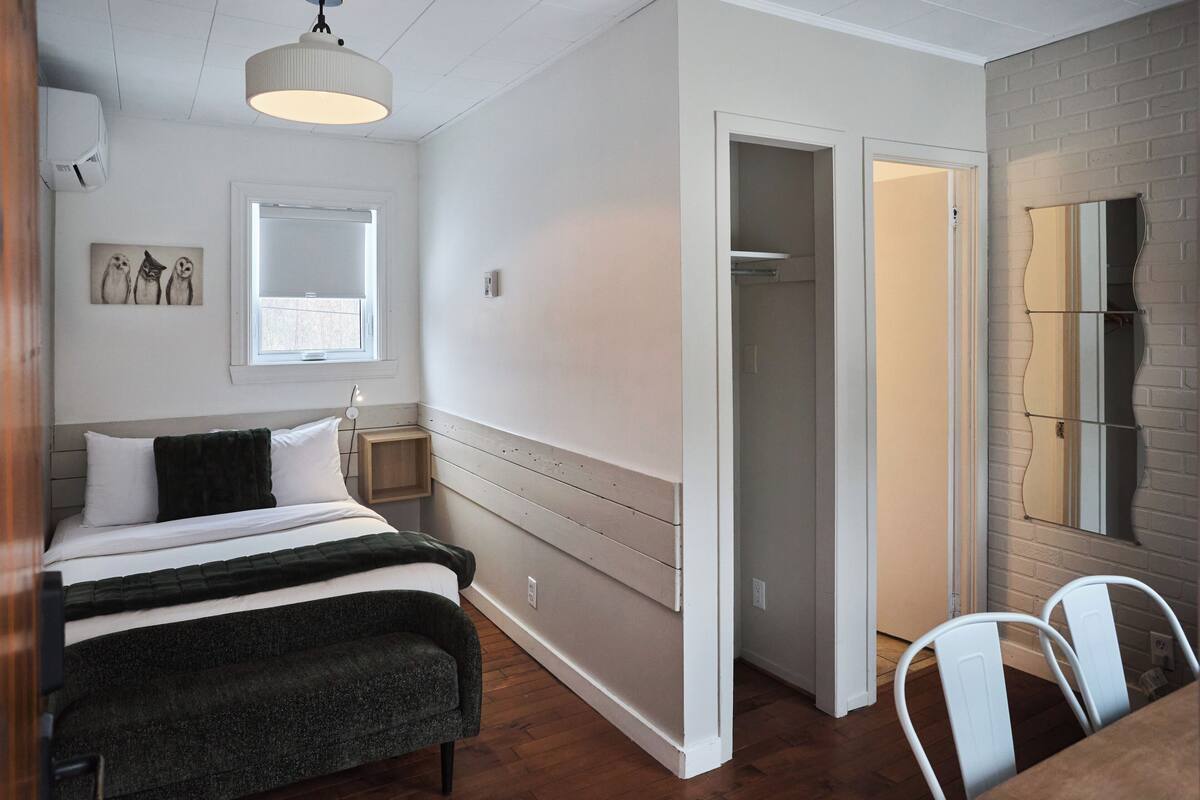 Quarto com cama, mesa de cabeceira e janela. Uma porta leva a outro cômodo.