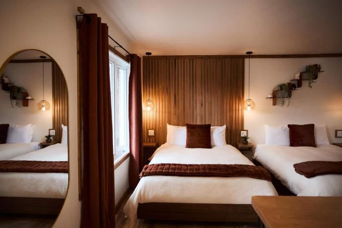 Quarto de hotel com duas camas, cabeceira de madeira, um espelho grande e uma janela com cortinas.