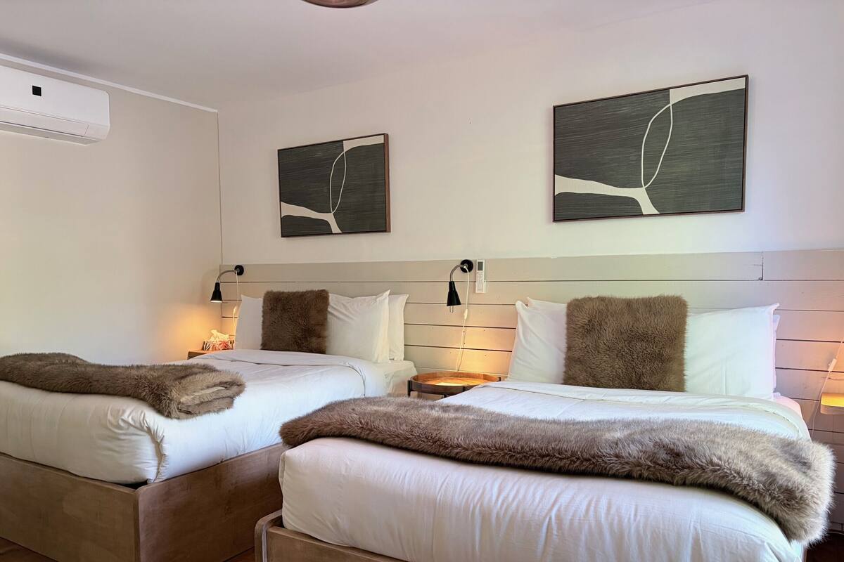 Quarto de hotel com duas camas, cada uma com colchão branco e manta marrom felpuda. Há obras de arte nas paredes acima das camas, ar condicionado no teto e abajures laterais.