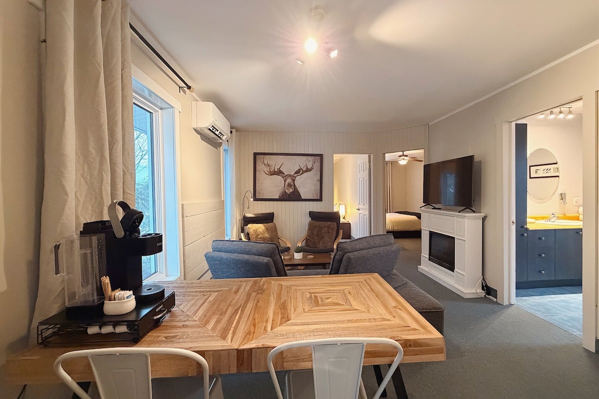 Uma sala de estar moderna com uma mesa de jantar de madeira, duas poltronas, um sofá, lareira e televisão.