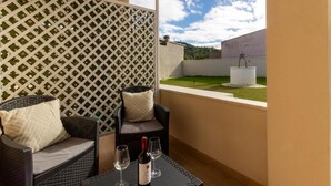 Uma varanda com móveis de vime, uma mesa com copos de vinho e vista para um gramado verde e um edifício.
