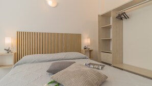 Quarto moderno com cabeceira de madeira, cama com cobertura acolchoada, duas almofadas, mesa de cabeceira com abajur e guarda-roupa de madeira.