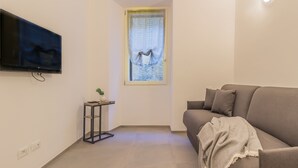 Uma sala de estar com televisão de tela plana na parede, uma janela com vista, um sofá e uma mesinha com uma planta.