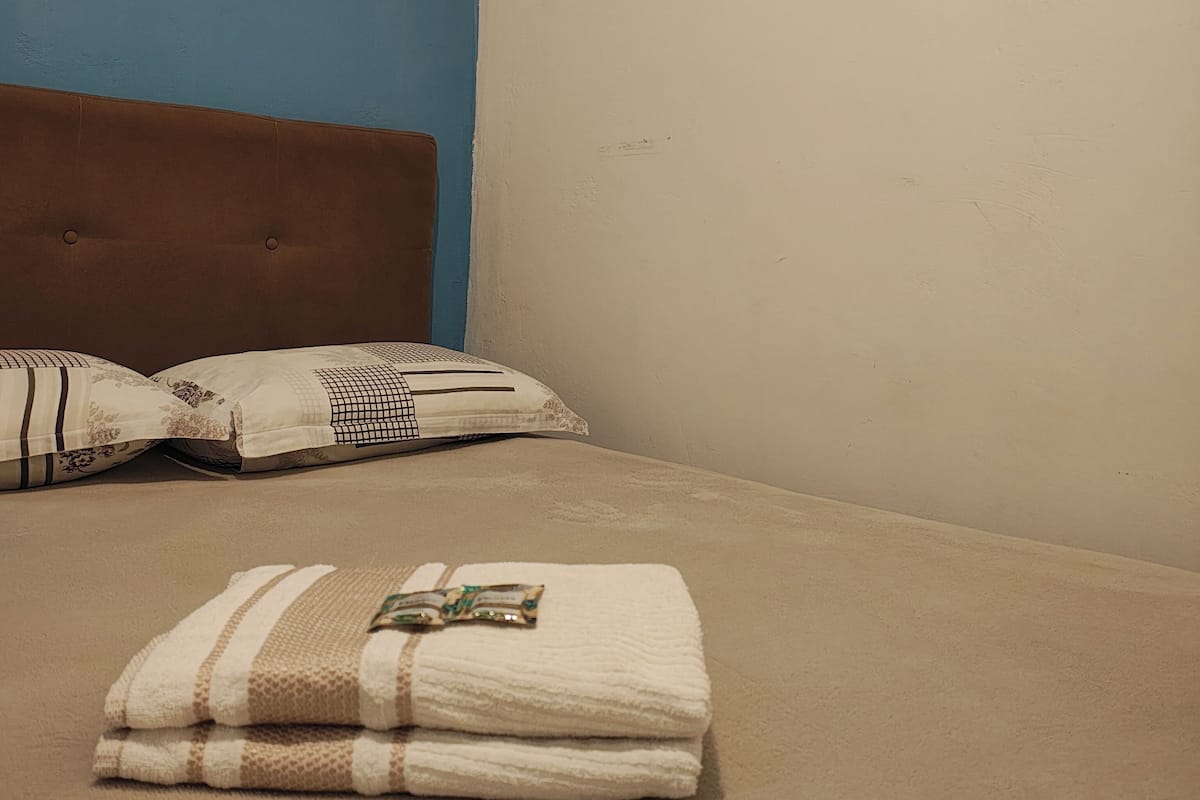 Uma cama com cabeceira marrom, duas almofadas estampadas e uma toalha dobrada com etiqueta.
