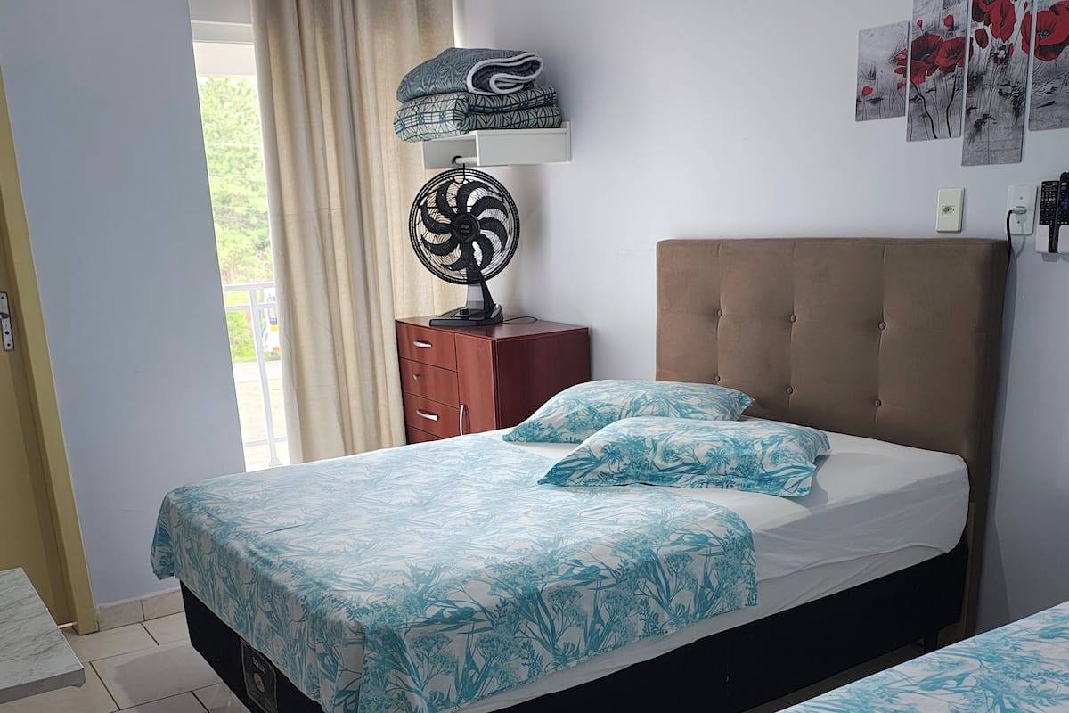 Quarto com cama, mesa de cabeceira, abajur e ventoinha fixada na parede.