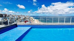 Uma piscina no terraço com vista para a cidade, céu claro e uma praia distante.