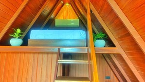 Quarto sótão triangular com uma cama, duas plantas em vasos e uma escada.