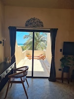 Quarto com porta de vidro deslizante que dá para uma varanda com vista para palmeiras e o oceano. Há uma cadeira de madeira e uma pequena mesa com um abajur.