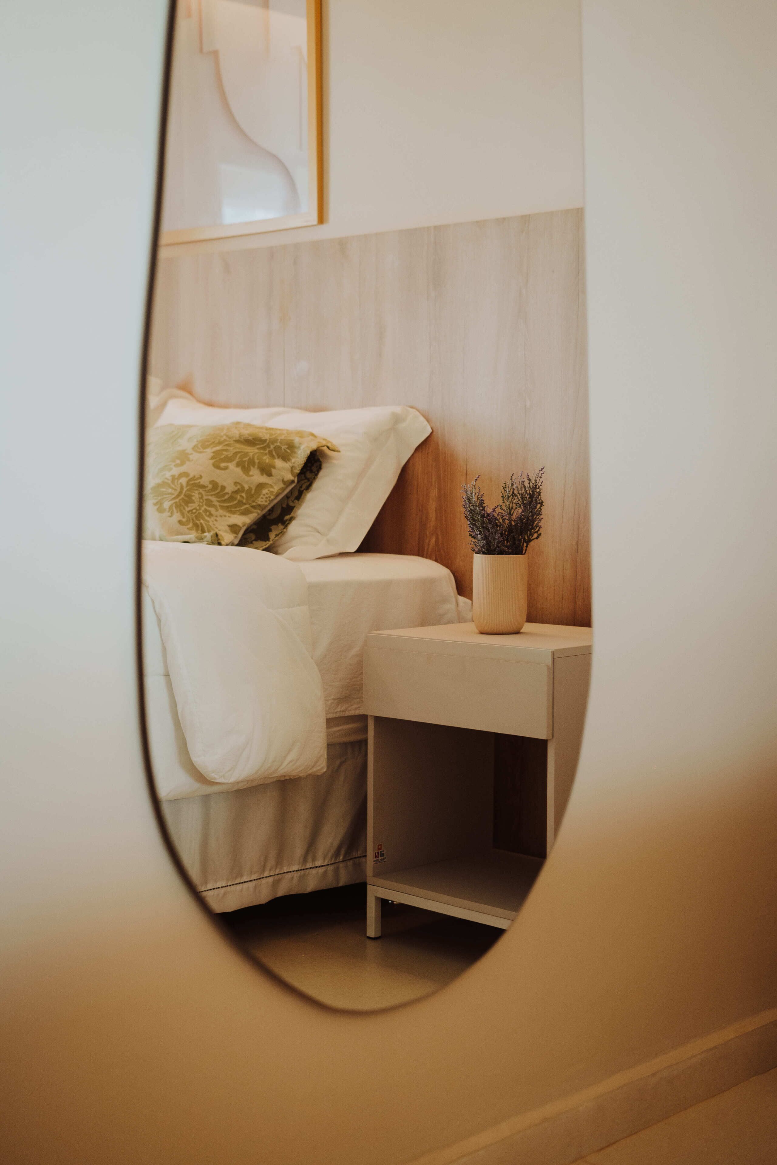 Quarto com cama, mesa de cabeceira e espelho refletindo parte do espaço.