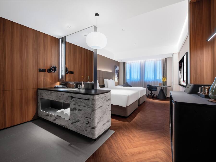 Quarto de hotel moderno com toucador em mármore, cama com roupa de cama branca, escrivaninha com cadeira e uma grande janela com cortinas.