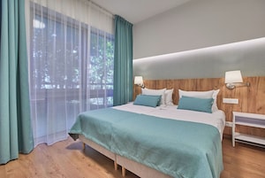 Quarto com cama, duas luminárias de cabeceira, mesa de apoio, janela com cortinas leves e vista para o verde lá fora.