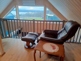 Uma varanda com vista para um lago e montanhas, uma poltrona de couro marrom, uma mesa pequena com um jogo americano floral e piso de madeira.