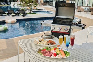 Uma área de refeições junto à piscina, com churrasqueira, prato de frutos do mar e bebidas.