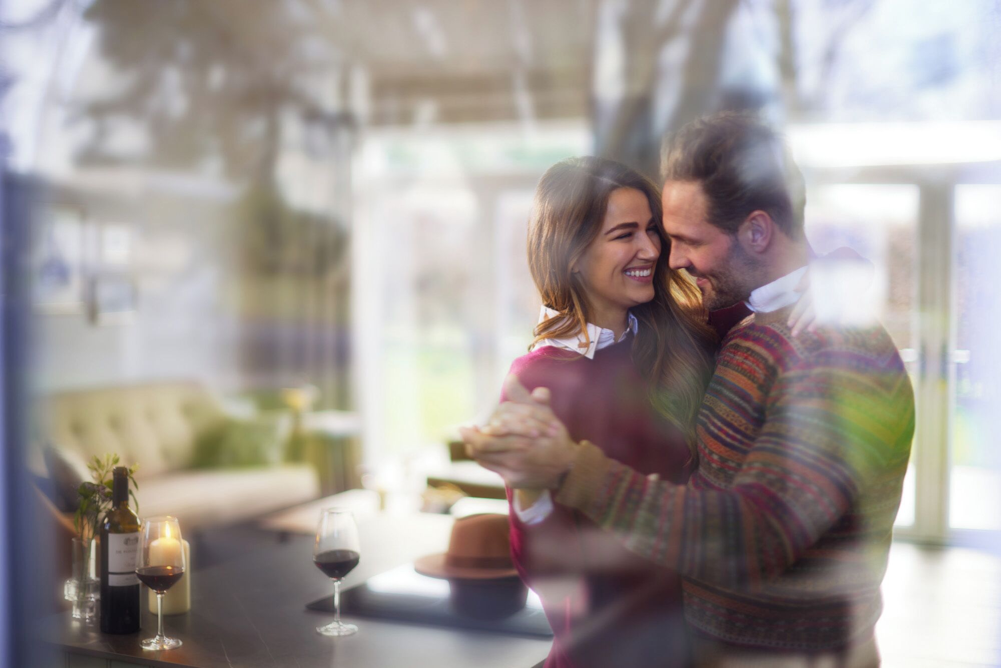 Um casal se abraçando à mesa, com taças de vinho e uma vela, em um quarto com uma grande janela e um sofá.