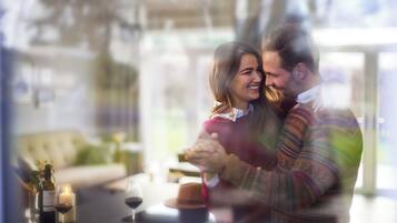 Um casal se abraçando à mesa, com taças de vinho e uma vela, em um quarto com uma grande janela e um sofá.