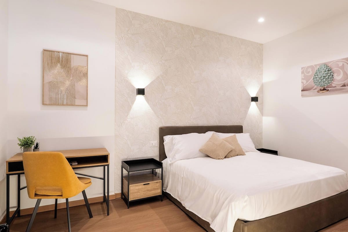 Quarto com cama, mesas de cabeceira, escrivaninha, cadeira e iluminação embutida na parede.