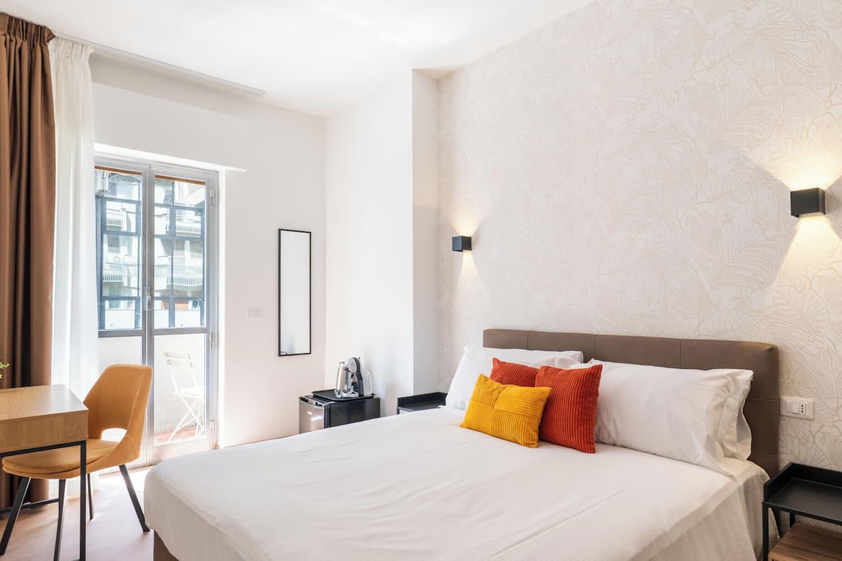 Quarto de hotel com cama, escrivaninha, cadeira, mesa de cabeceira e luminária na parede.