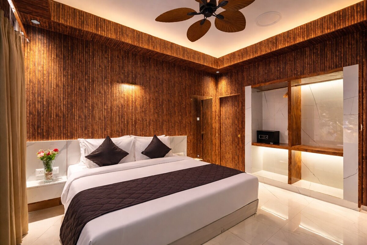 Um quarto moderno com parede de madeira, uma cama com roupa de cama branca e almofadas escuras, uma mesa de cabeceira com um vaso de flores e um ventilador de teto.