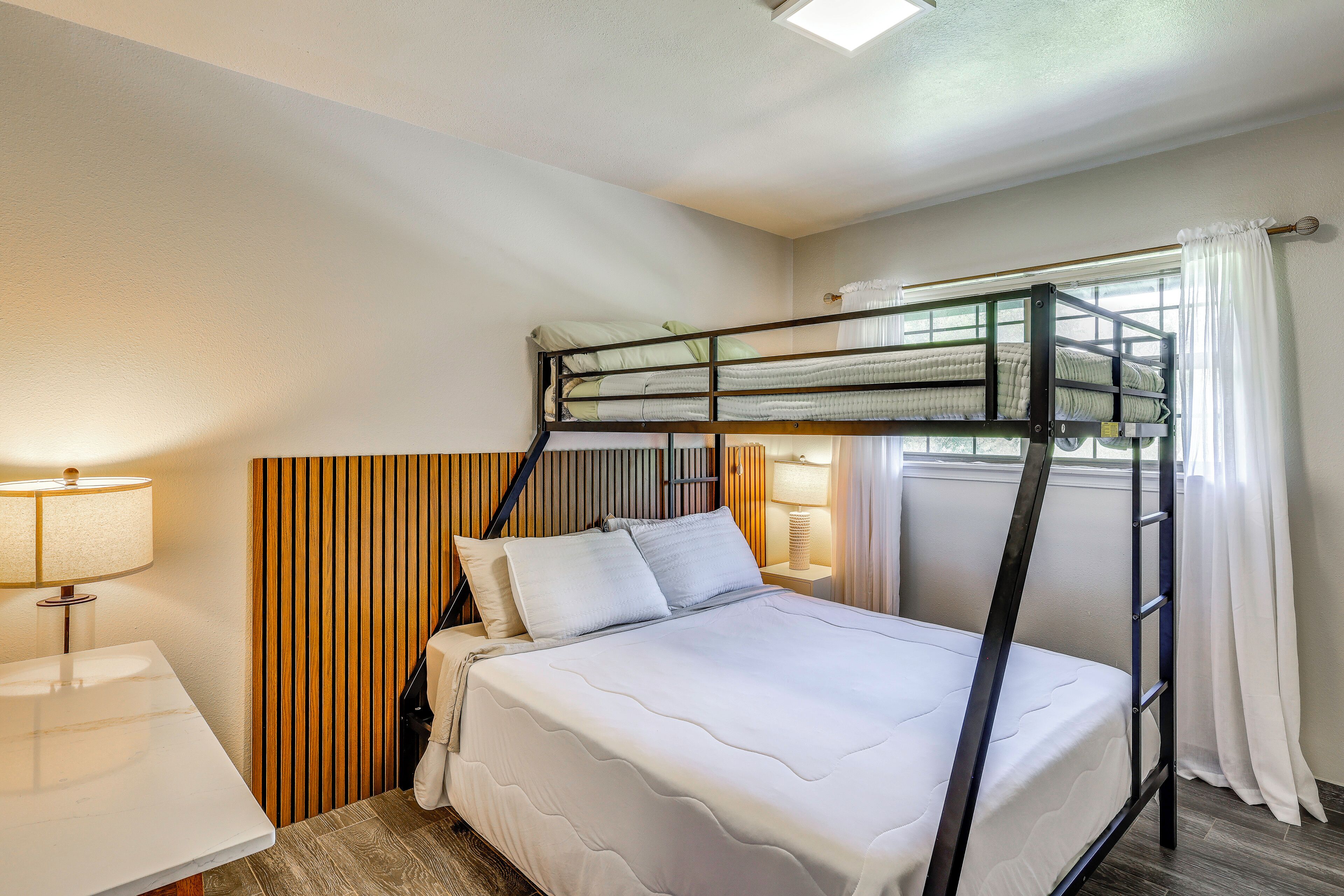 Uma cama beliche com escada, mesa de cabeceira, abajur e janela com cortinas.