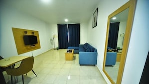 Quarto com televisão na parede, sofá azul, mesa de centro de madeira, escrivaninha com cadeira e um espelho grande.