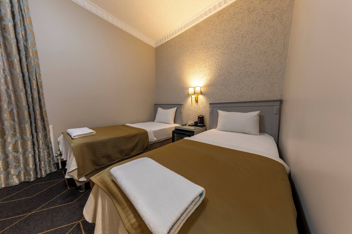 Quarto de hotel com duas camas, uma mesinha de cabeceira, uma luminária e uma cortina.