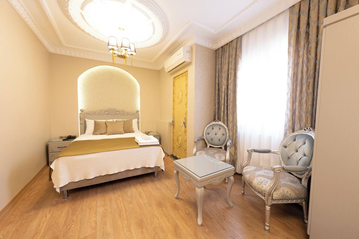 Quarto de hotel com cama, mesa, duas cadeiras, um candelabro e uma janela grande com cortinas.
