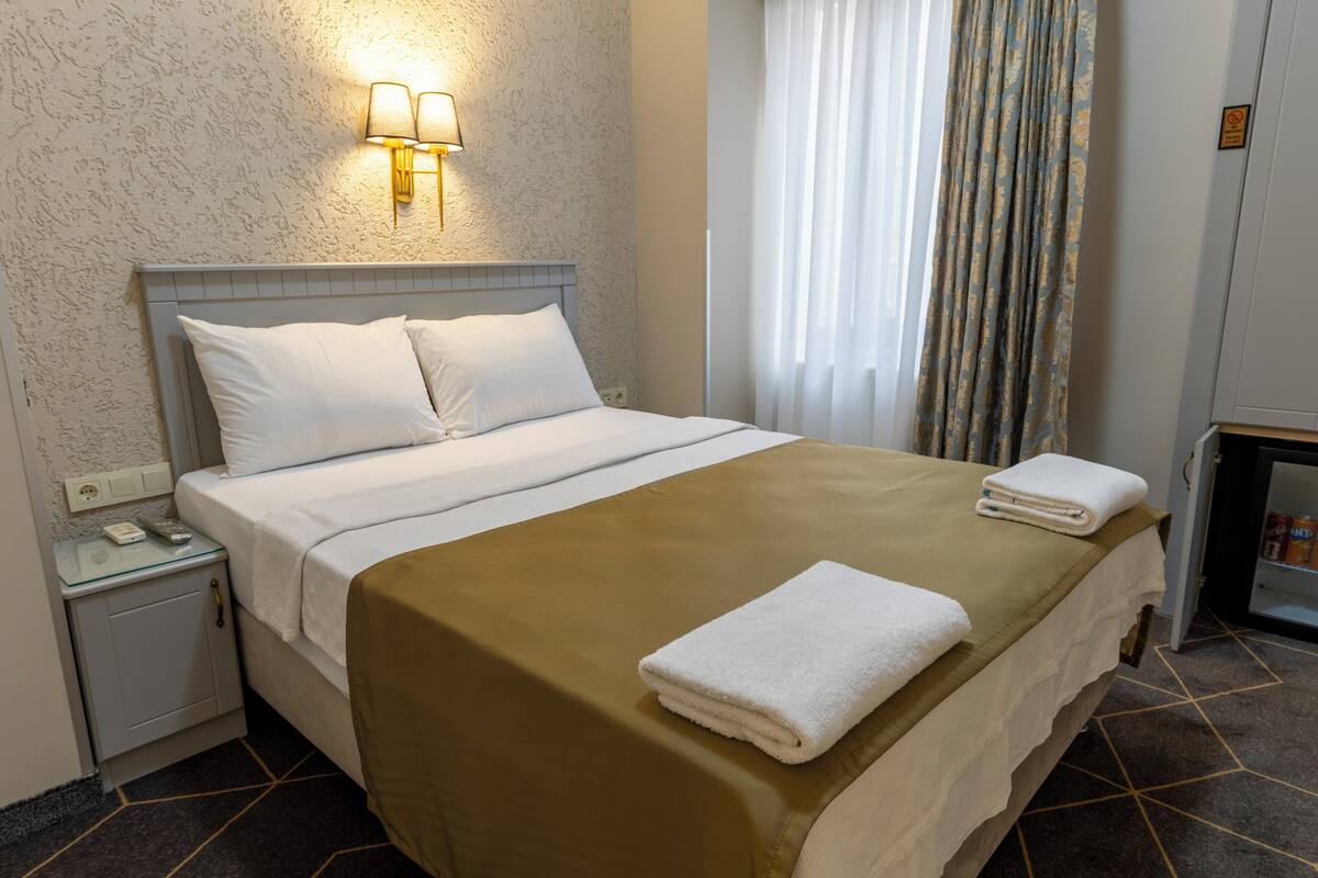 Quarto de hotel com cama, luminárias de cabeceira, mesa de cabeceira, janela com cortinas e minibar.