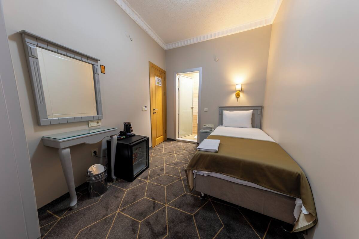 Quarto de hotel com uma cama, escrivaninha, espelho, mesa de cabeceira e porta que dá para outro quarto.